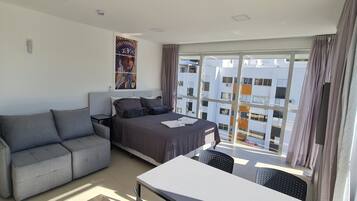 Quarto de hotel moderno com cama, sofá cinzento e área de refeições.