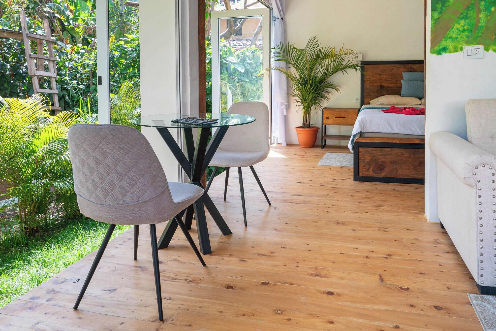 Um quarto aconchegante com piso de madeira, uma cama com encosto, uma mesinha com cadeiras e um vaso de planta.