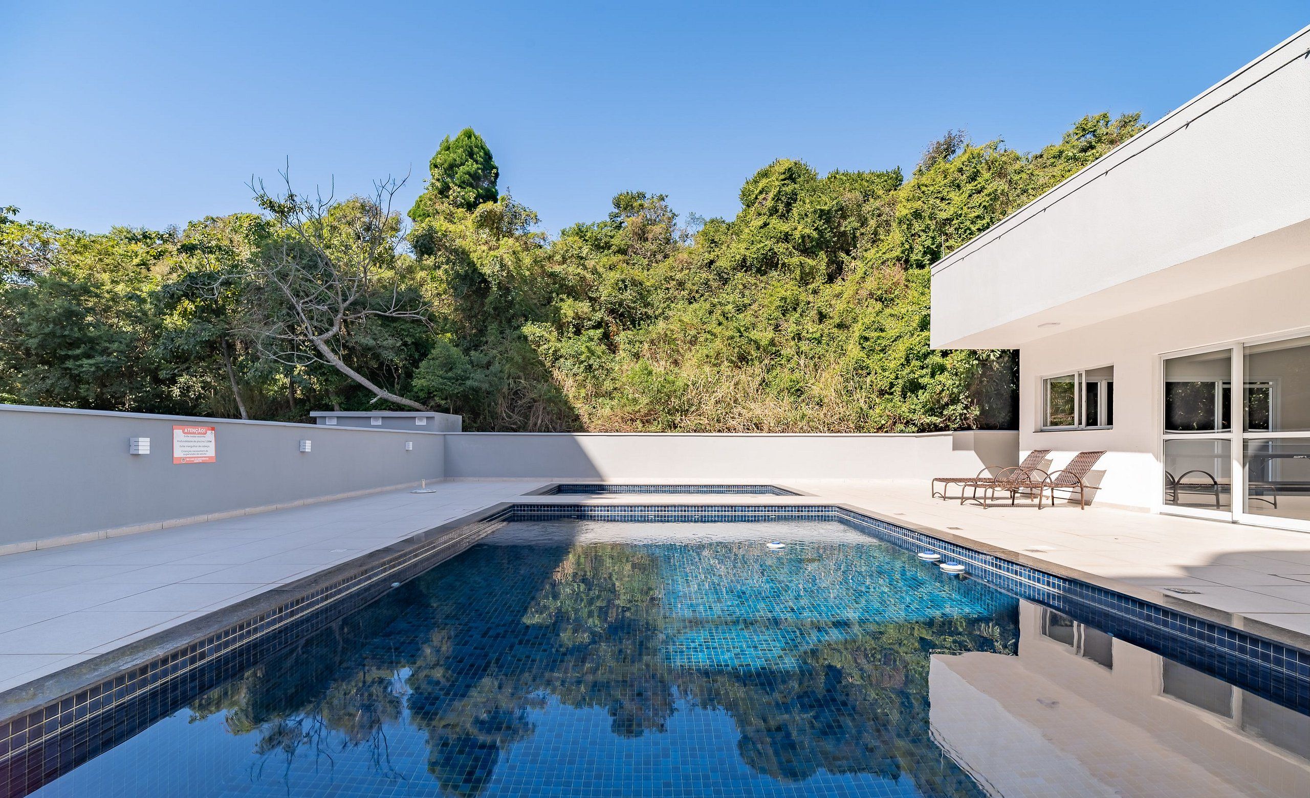 Piscina com água azul cristalina, rodeada por um edifício moderno e vegetação exuberante.