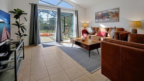 Uma sala de estar espaçosa com um sofá grande, uma mesa de centro e uma televisão. Há uma porta de vidro que dá para uma área externa com piscina.