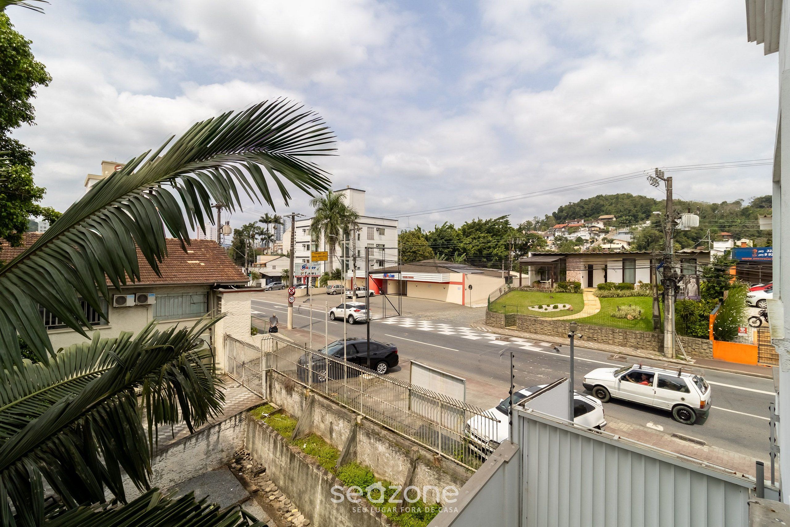Vista da rua com um carro branco na estrada, uma faixa de pedestres e um edifício com telhado vermelho ao fundo.