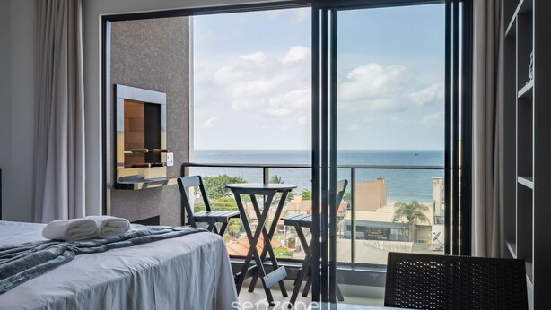 Quarto de hotel com uma varanda ampla, uma cama bem arrumada, uma pequena mesa e cadeiras, e vista para o mar.
