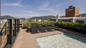 Área de piscina no terraço, com cadeiras de descanso e vista para a cidade e colinas.