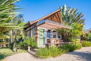 Uma cabana com porta de vidro, rodeada por plantas tropicais e um caminho de cascalho.