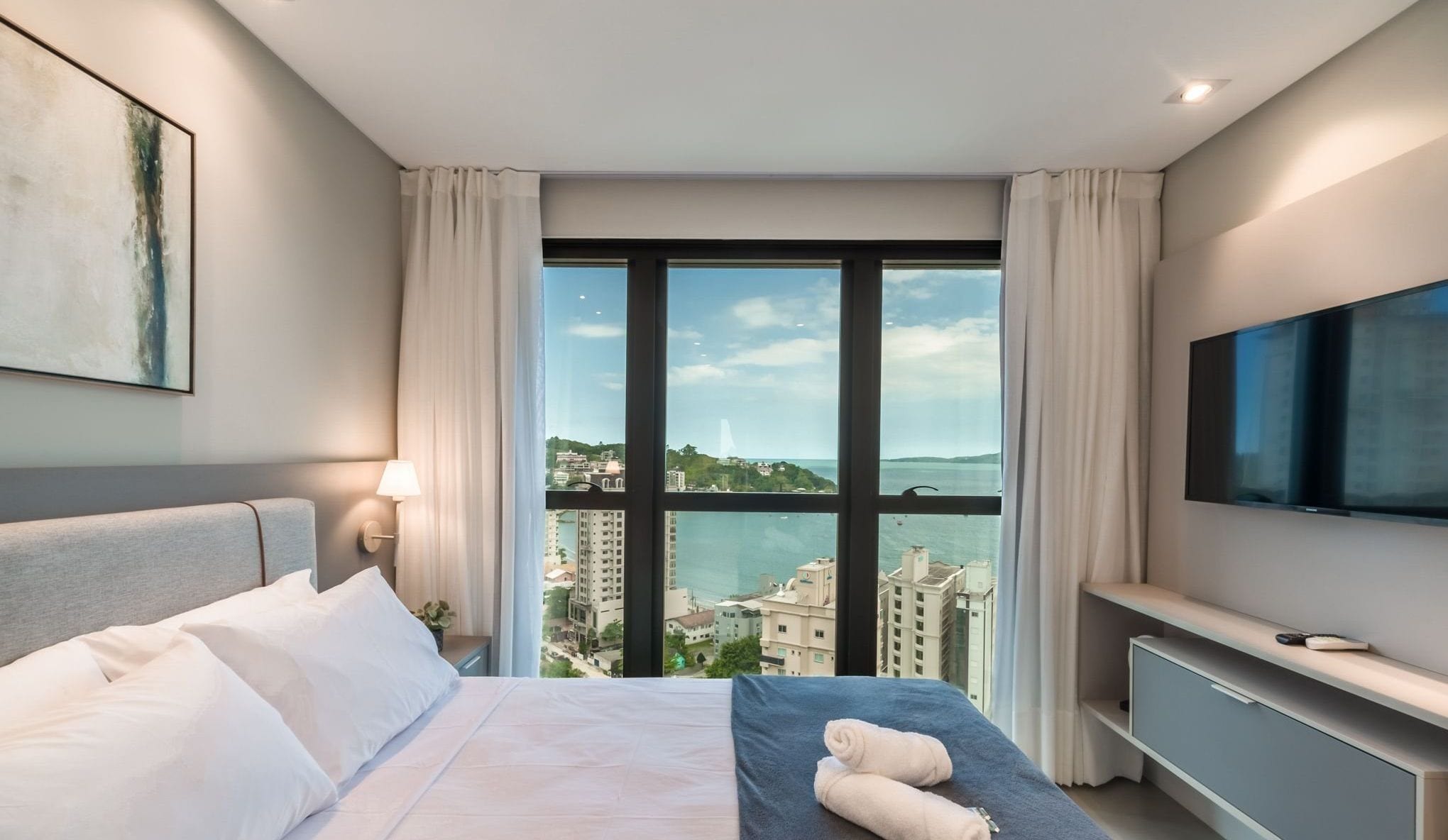 Quarto de hotel com cama, televisão, uma grande janela com vista para a cidade e para a água, e um quadro na parede.