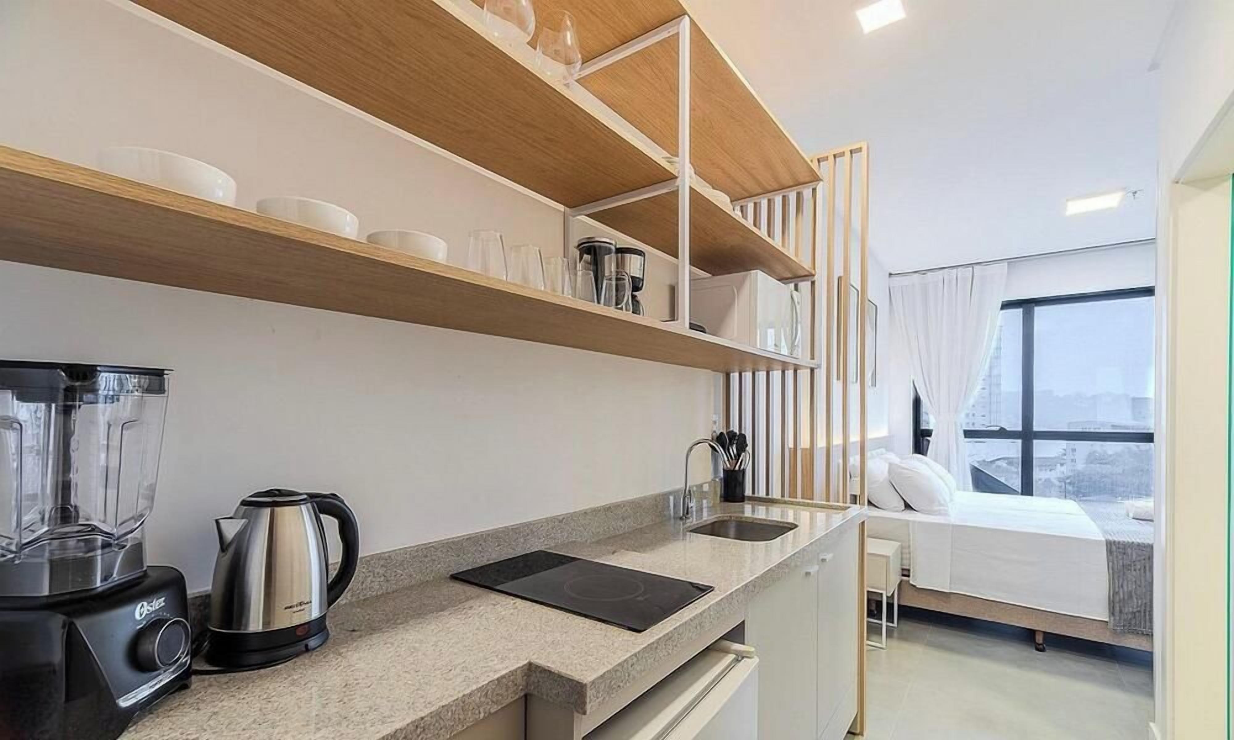 Cozinha moderna com máquina de café integrada, chaleira e liquidificador. Há uma cama com vista para a cidade ao fundo.