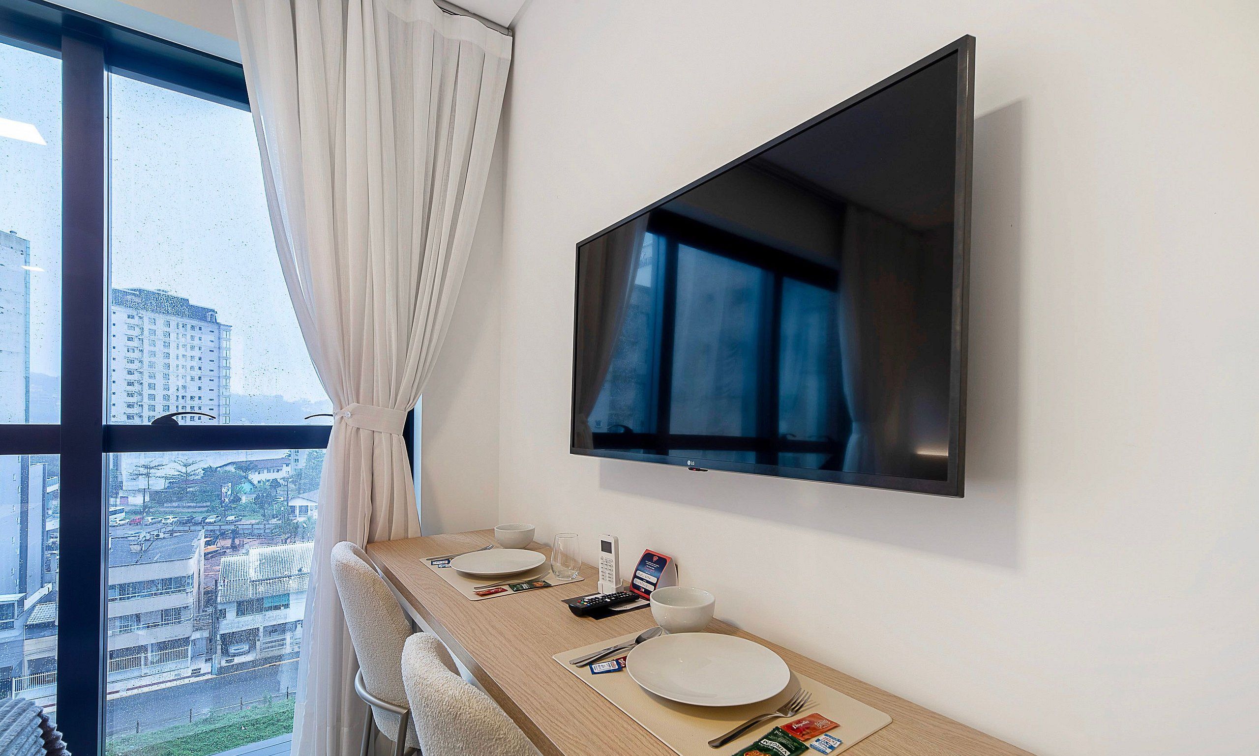 Quarto de hotel com uma grande janela, uma TV de tela plana na parede, uma mesa de madeira com um controle remoto, uma tigela e um copo.