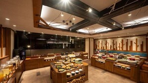 Uma área de pequeno-almoço tipo buffet com diversas opções de comida e bebidas, mobiliário de madeira e um design de teto moderno.