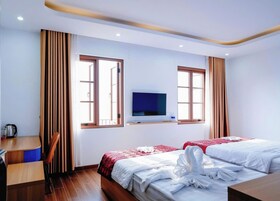 Quarto de hotel com duas camas, uma mesa, uma cadeira, televisão e janelas amplas com cortinas.