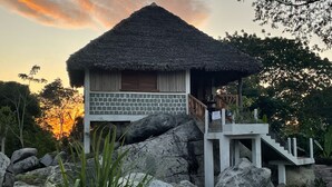 Uma casa com telhado de palha, fachada de pedra, um deque de madeira e uma escadaria que leva a um alpendre.