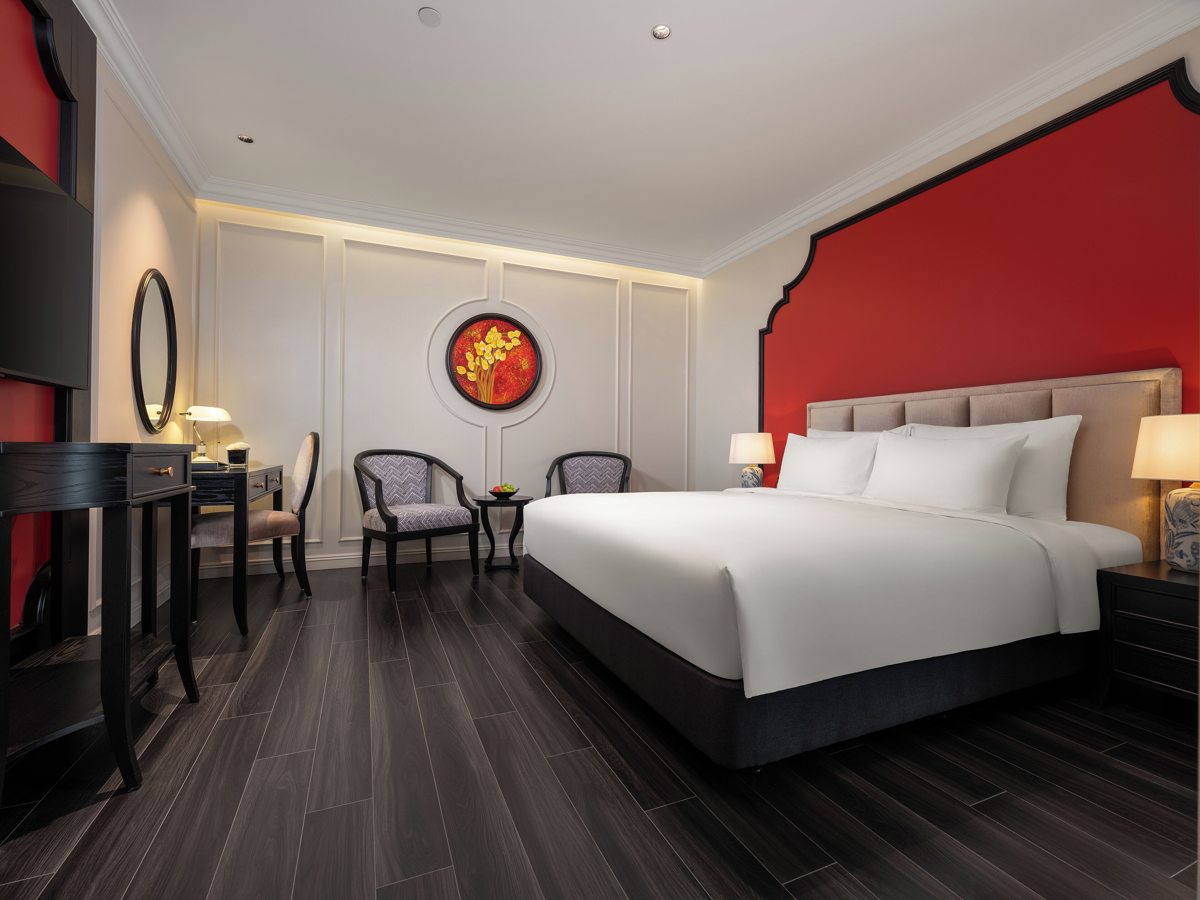 Quarto de hotel com uma cama grande, duas cadeiras, uma escrivaninha e uma parede de destaque vermelha.