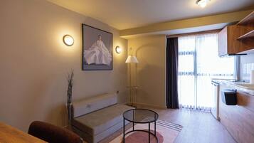 Quarto de hotel moderno com sofá, mesa redonda pequena, luminária, quadro na parede e vista para o exterior pela janela.