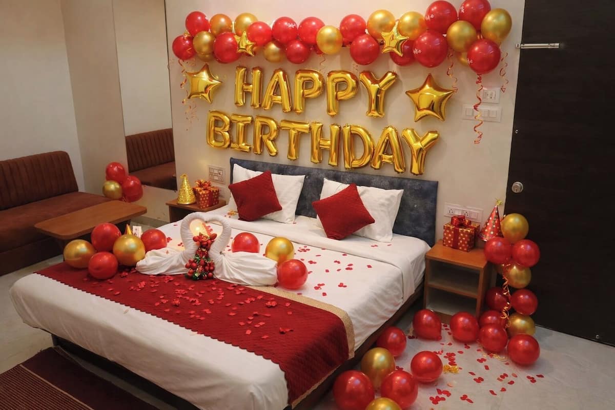Quarto de hotel decorado para um aniversário, com uma cama, almofadas e balões com a mensagem 'HAPPY BIRTHDAY'.
