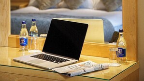 Quarto de hotel com cama, um laptop e jornal na mesa.