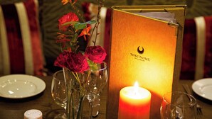 Mesa iluminada por velas, com um menu, um vaso de rosas vermelhas e um saleiro e pimenteiro.