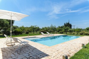 Piscina retangular com deck ladrilhado, rodeada por espreguiçadeiras e um guarda-sol branco.