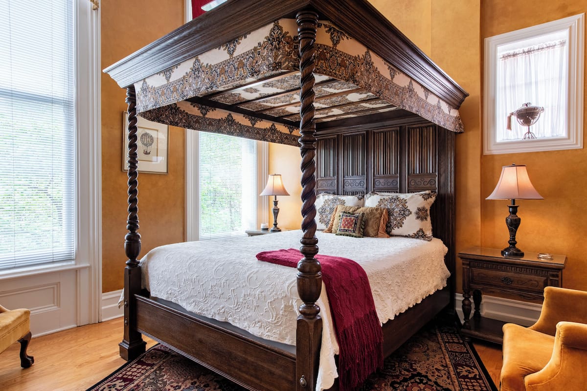 Uma cama com dossel e estrutura de madeira com detalhes entalhados, um cobertor vermelho e um cabeceira combinando. Há duas mesas de cabeceira com abajures, uma cadeira e um tapete estampado no assoalho de madeira.