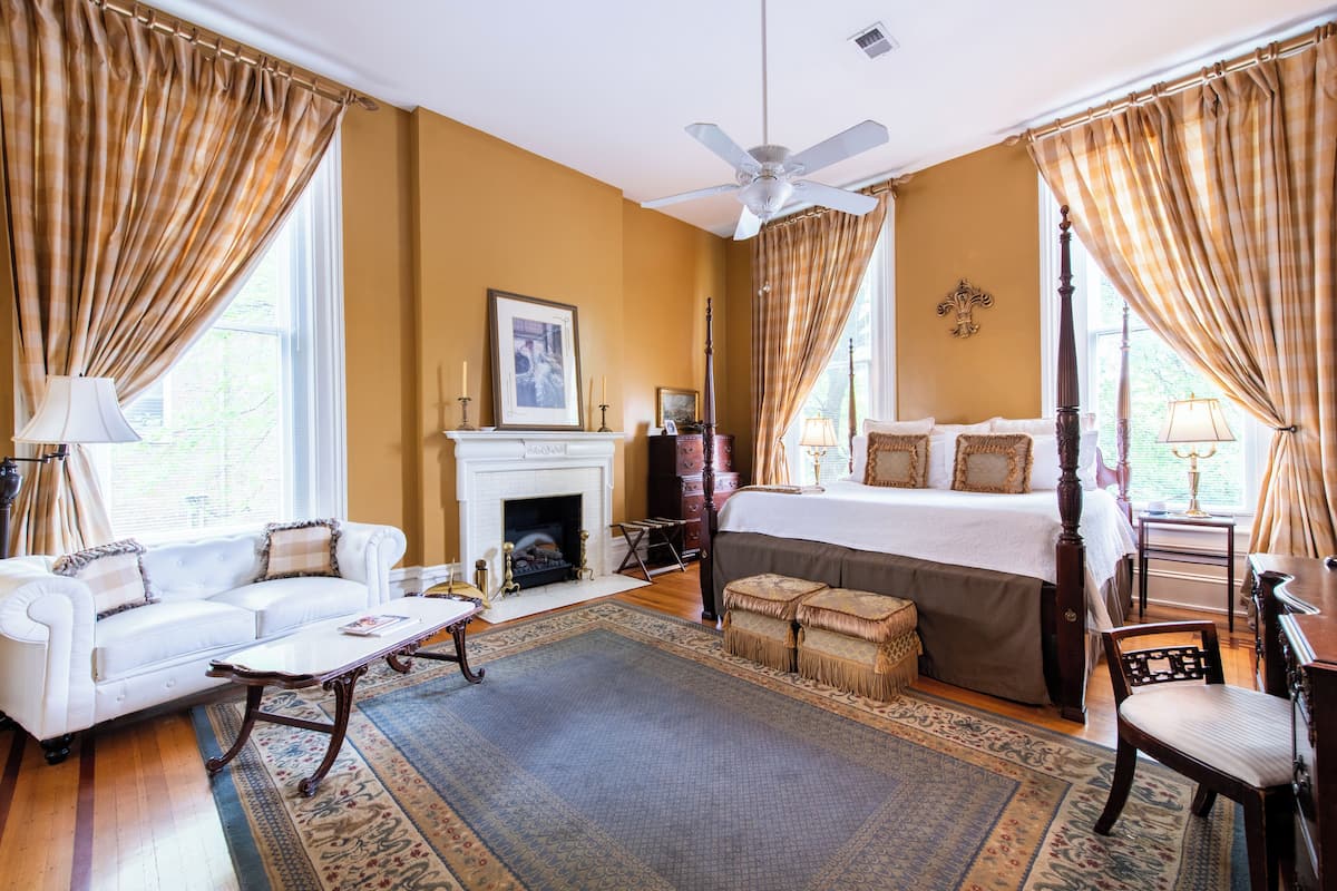 Quarto com sofá branco, mesa de centro de madeira, cama grande com cabeceira de madeira, lareira e ventilador de teto.