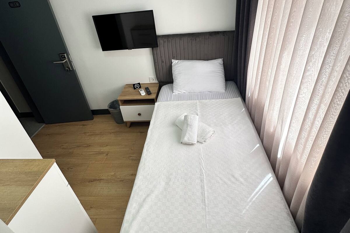 Uma cama de solteiro com colcha branca, uma televisão de tela plana na parede, uma mesa de cabeceira de madeira com controle remoto, um guarda-roupa cinza e uma janela com cortinas leves.