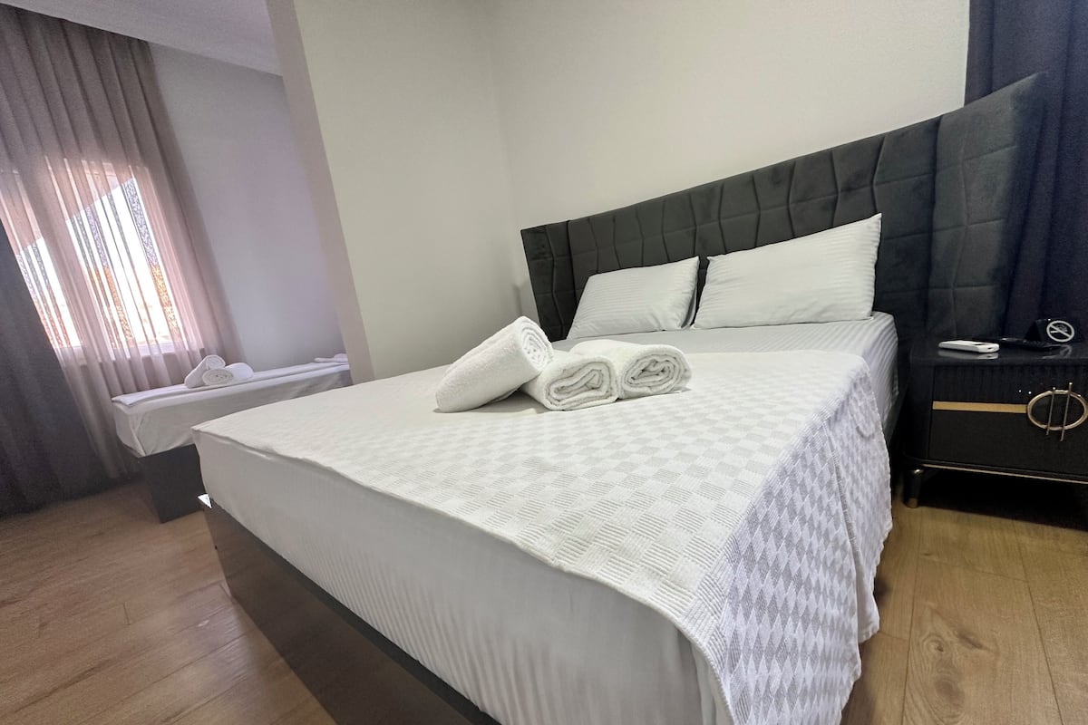 Cama bem arrumada com lençóis brancos e toalhas dobradas, cabeceira escura e mesa de cabeceira com telefone e um pequeno objeto.