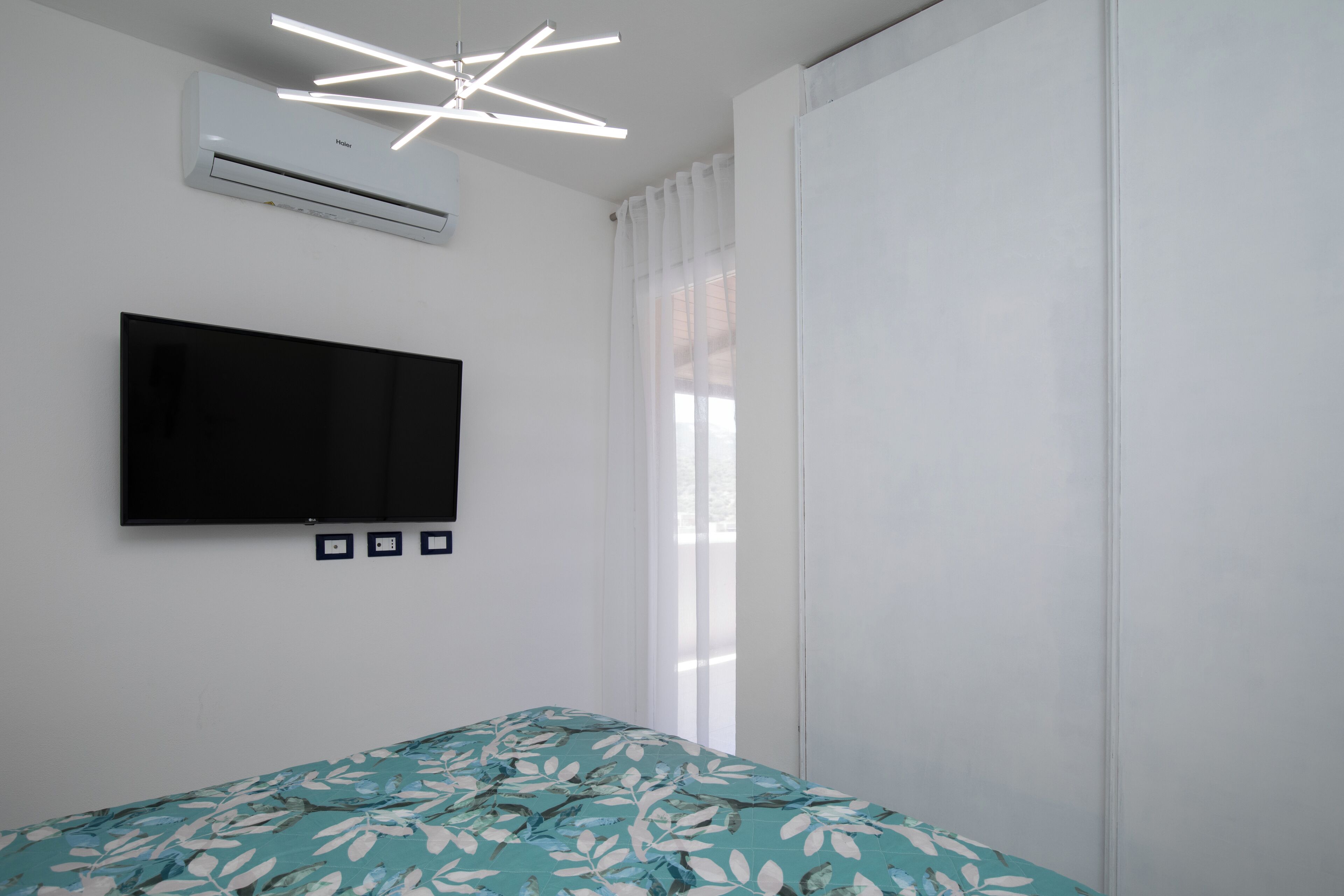 Quarto com cama, TV na parede, ar condicionado e janela com cortinas.