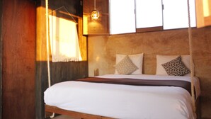 Uma cama com estrutura de madeira, roupa de cama branca e almofadas estampadas, luminária pendente e janela com vista.