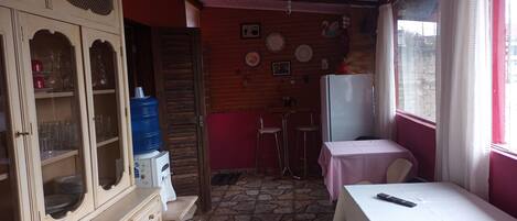 Um quarto pequeno e acolhedor com piso de azulejos, uma mesa branca com toalha, um refrigerador, um armário de madeira com portas de vidro e uma janela com cortinas.
