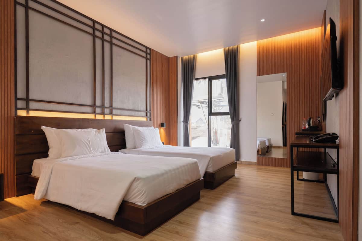 Quarto de hotel com duas camas, piso de madeira, uma janela grande com cortinas, parede de painéis de madeira e televisão na parede.