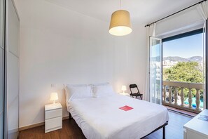 Quarto com cama, mesas de cabeceira, luminária, cadeira e varanda com vista para prédios e árvores.