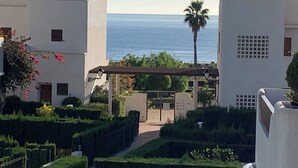 Zona residencial com edifícios brancos, jardim com sebes, uma pérgula e vista para o mar.