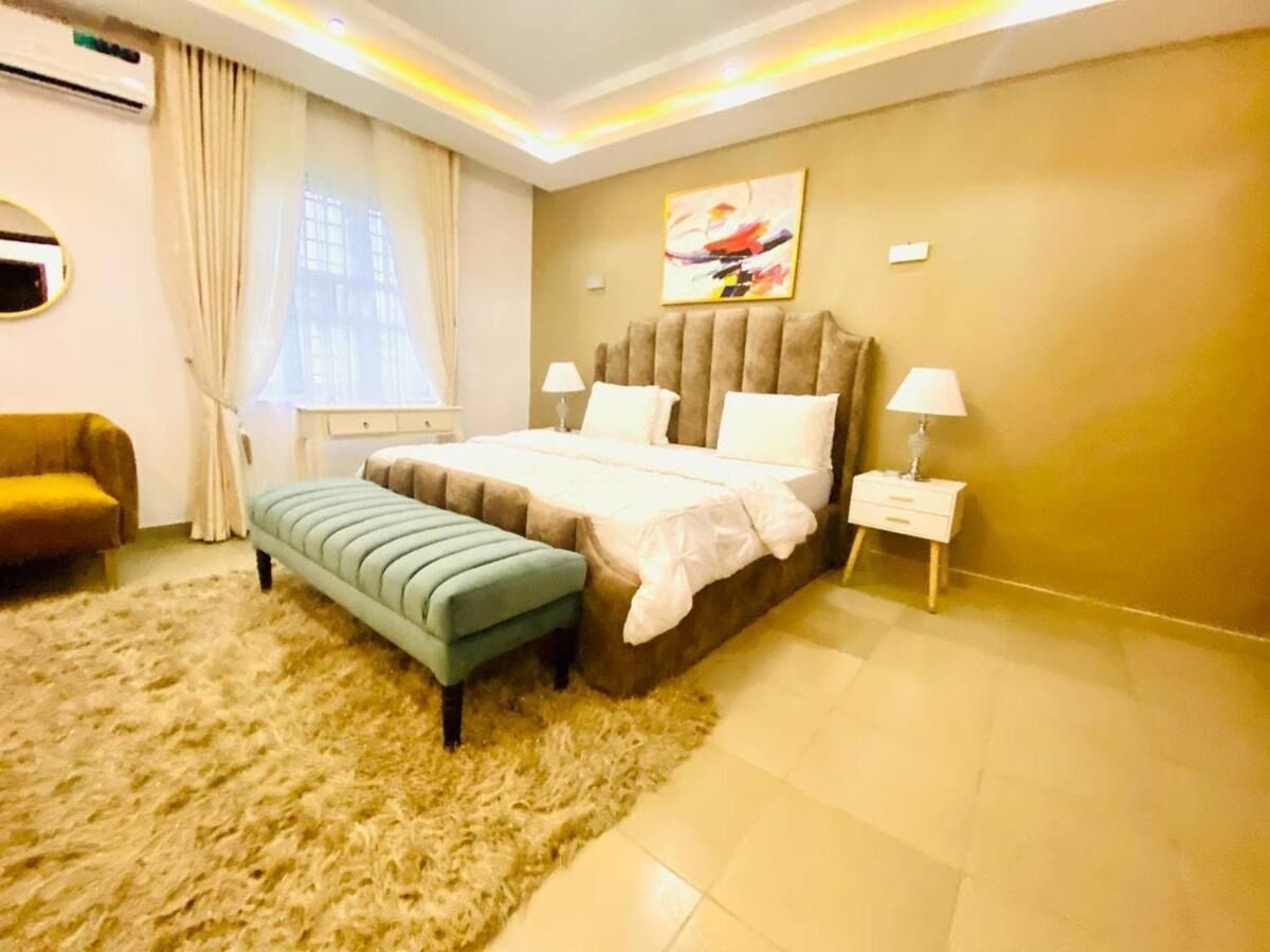 Quarto com cama grande, um banquinho, duas mesas de cabeceira com abajures, uma janela com cortinas e um quadro na parede.