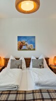 Quarto de hotel com duas camas, um quadro de uma vaca e um cão, e duas luminárias de cada lado.