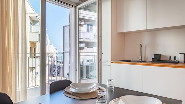 Espaço de refeições com mesa posta para quatro pessoas, área de cozinha com pia e bancada, e vista da cidade através de grandes janelas.