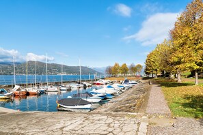 Uma marina com barcos atracados ao longo de um cais de pedra, um caminho gramado e árvores com folhas de outono.