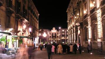 Vista noturna de uma praça histórica com edifícios ornamentados, uma fonte central e pessoas caminhando.