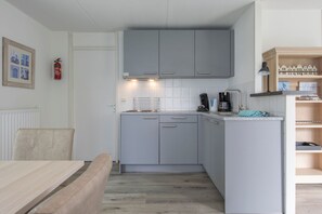 Uma cozinha compacta com armários cinza, bancada com chaleira e prateleira pequena com objetos decorativos.