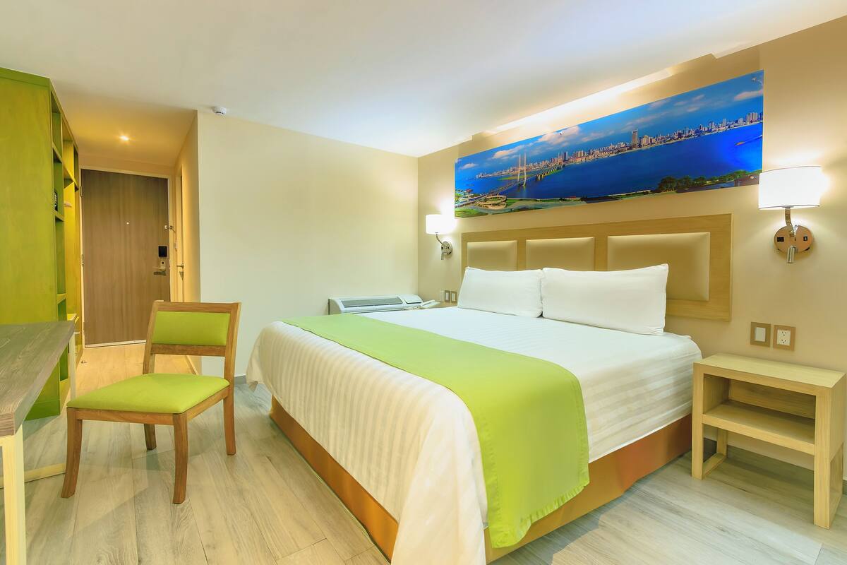 Quarto de hotel com uma cama grande, uma poltrona verde, uma mesinha de cabeceira e uma pintura de paisagem urbana na parede.