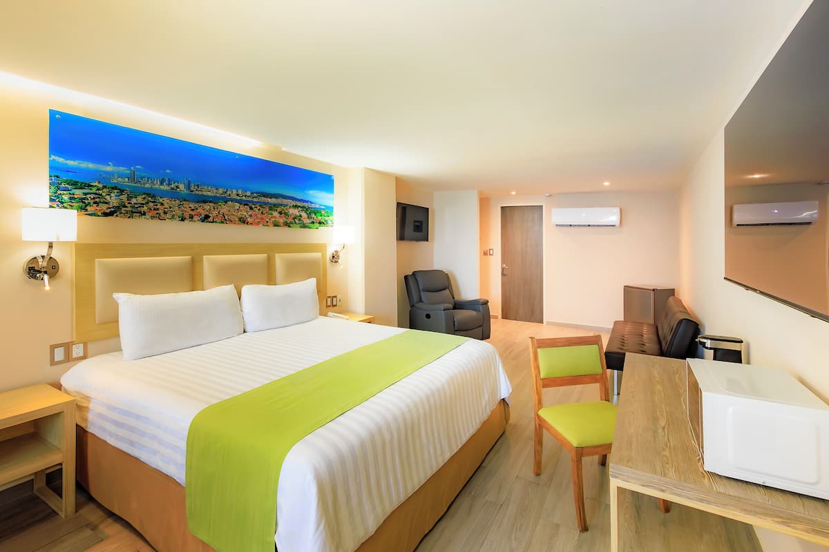 Quarto de hotel com uma cama grande, escrivaninha, cadeira, televisão e aparelho de ar condicionado.