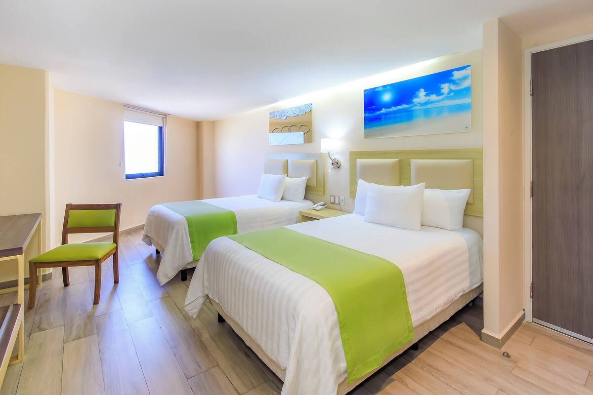Quarto de hotel com duas camas, uma escrivaninha, uma cadeira, uma janela e um quadro na parede.