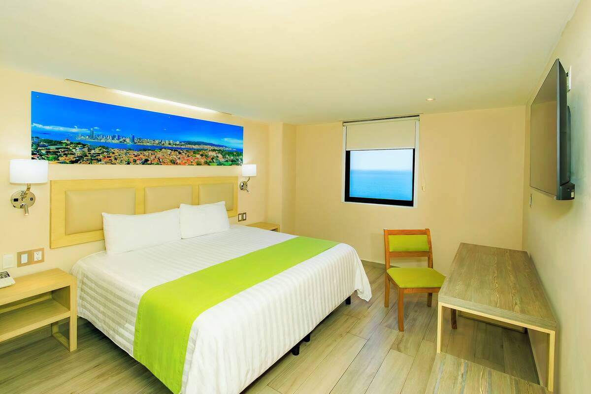 Quarto de hotel com cama, escrivaninha, cadeira, televisão e pintura de uma paisagem urbana.