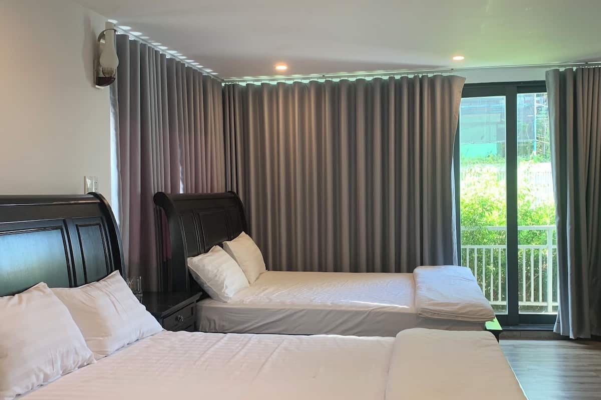 Quarto de hotel com duas camas, uma cabeceira escura e vista para o verde pela janela.