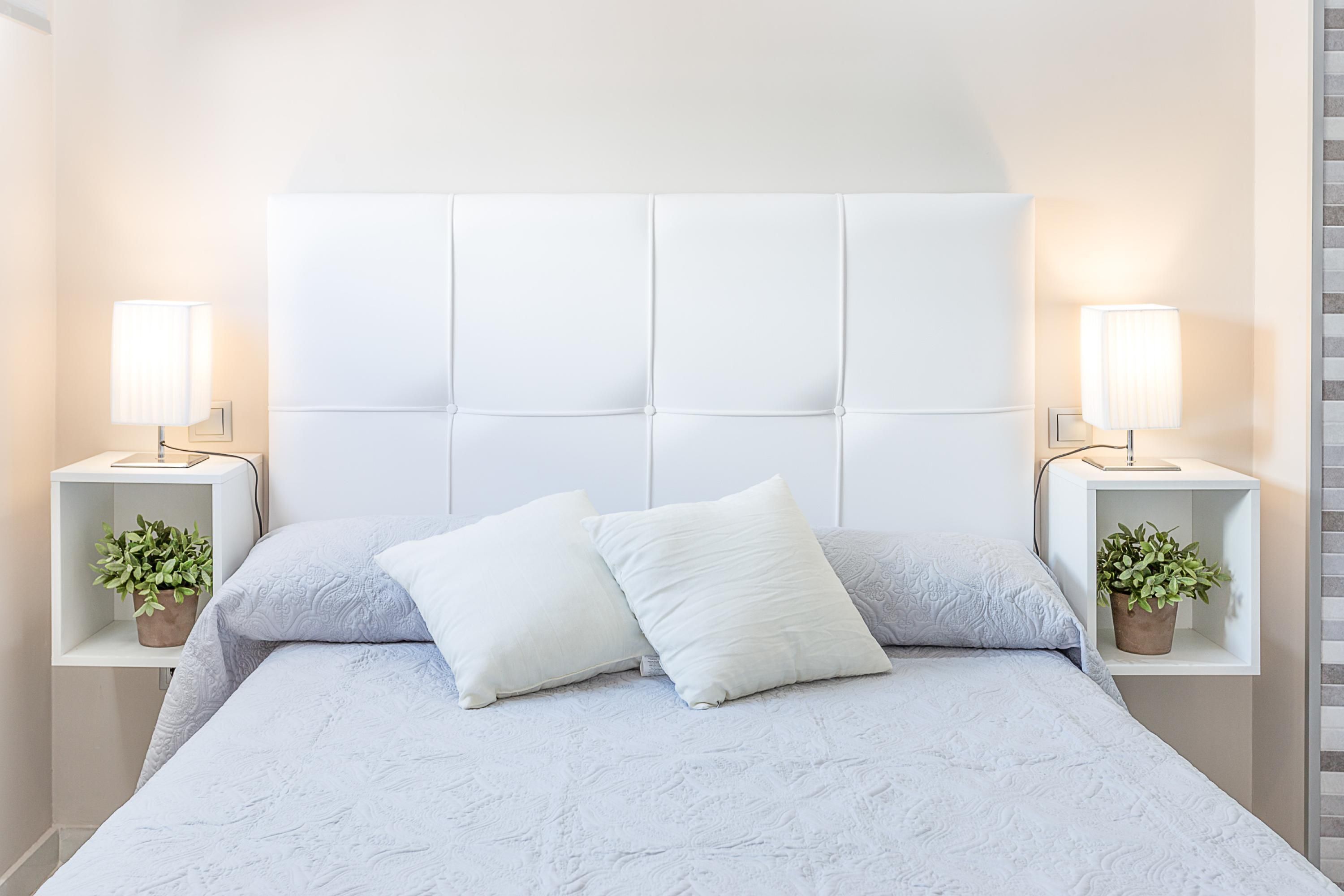 Cama bem arrumada com cabeceira estofada, duas mesas de cabeceira com luminárias e plantas em vasos, e uma parede em tom claro.