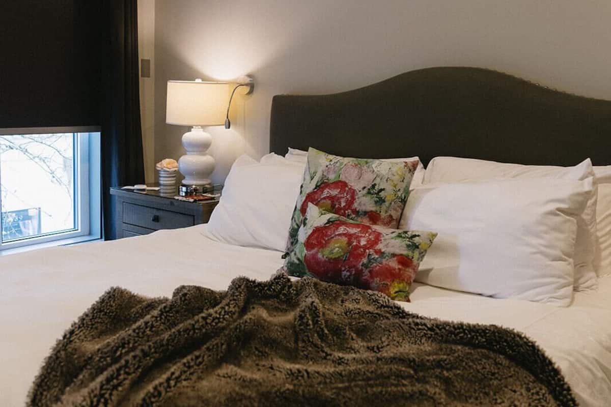 Cama bem arrumada com lençóis brancos, cabeceira escura e almofada floral. Há uma mesa de cabeceira com uma luminária e uma pequena pilha de livros. O quarto tem uma janela com vista para árvores e um prédio lá fora.