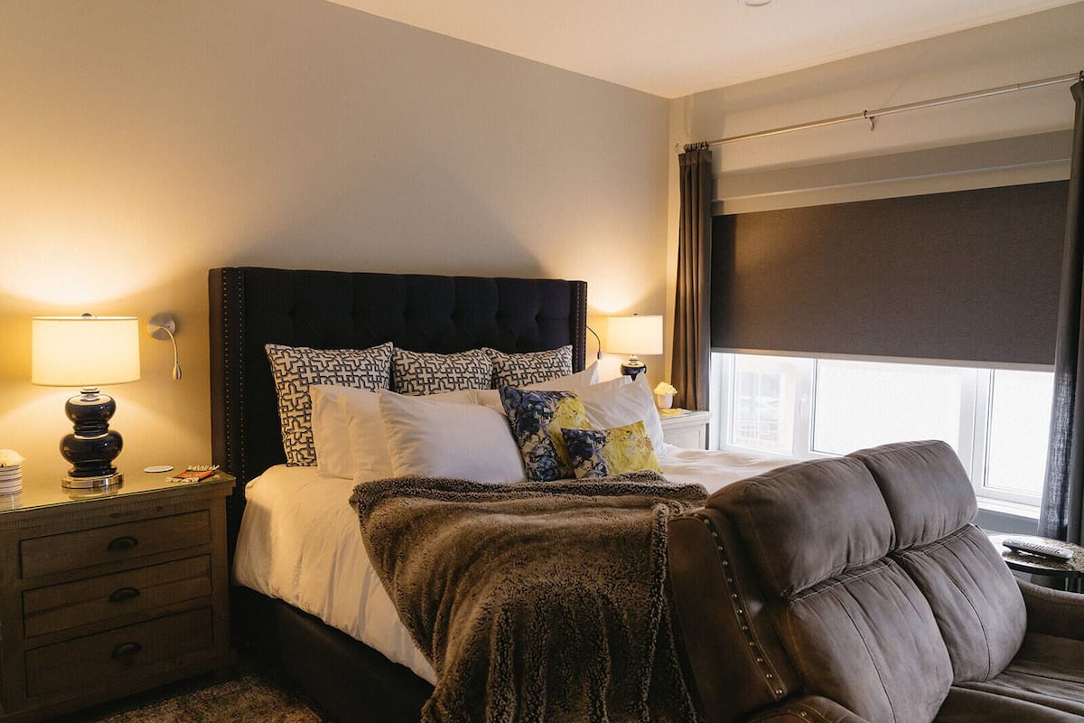 Quarto de hotel com uma cama grande, uma poltrona de couro, uma mesa de cabeceira com abajur e uma janela com cortinas.