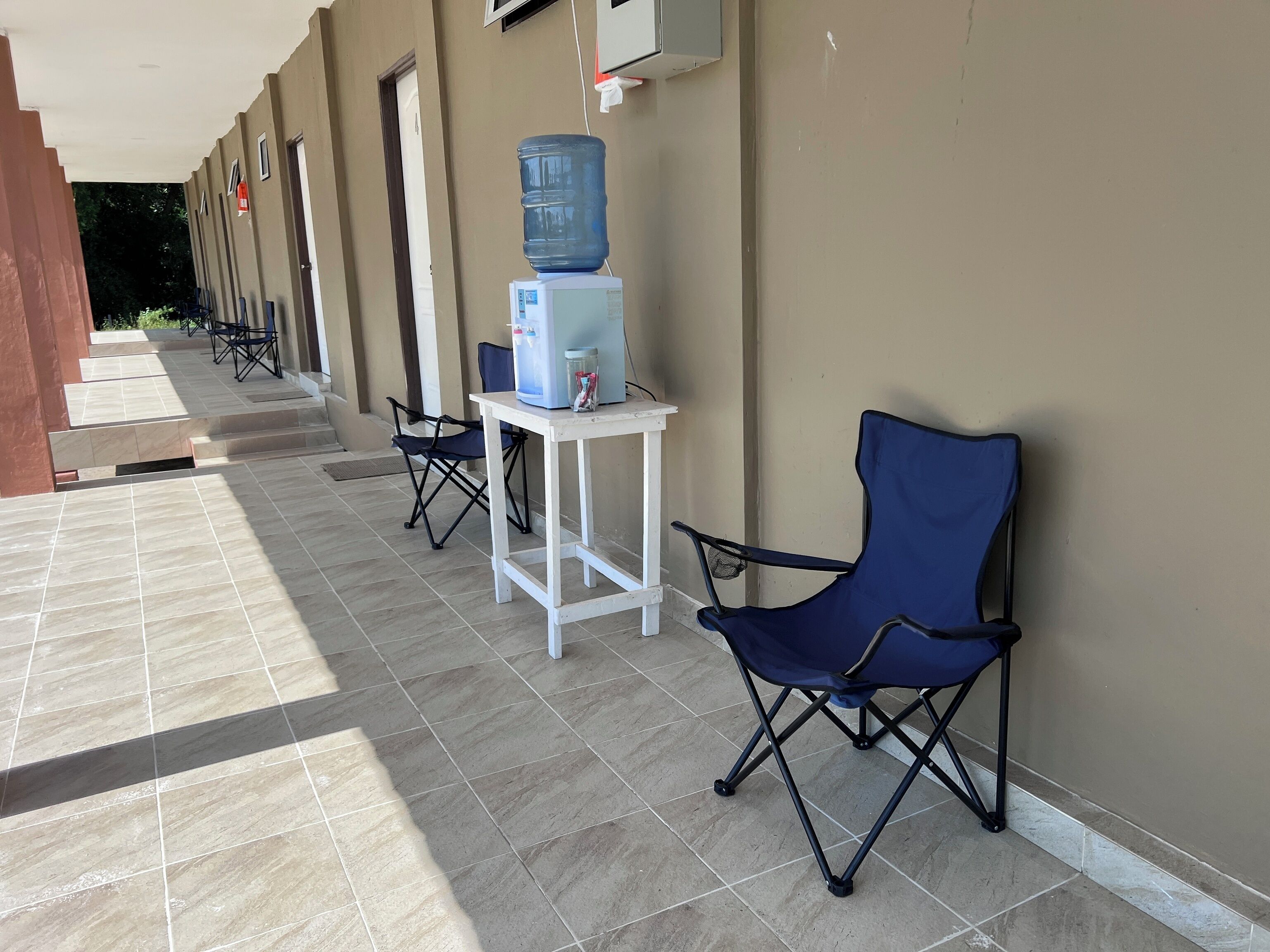 Área externa coberta com piso de azulejos, bebedouro, mesa e cadeiras azuis dobráveis.