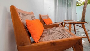 Uma cadeira de lounge de madeira com assento trançado e almofada laranja, posicionada em frente a uma parede branca com janela e porta.