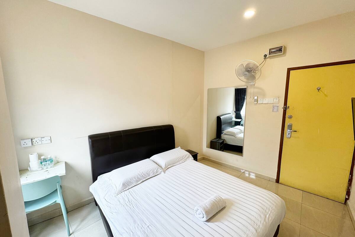 Quarto de hotel com cama, escrivaninha, espelho, ventilador e porta.