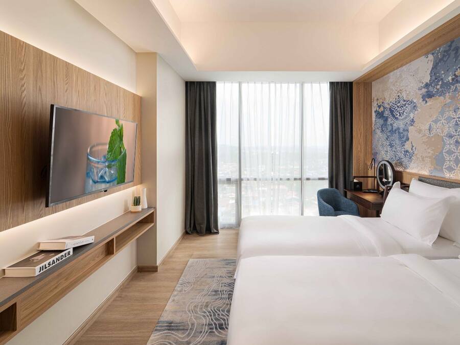 Quarto de hotel moderno com uma cama grande, televisão de tela plana, escrivaninha com abajur e vista para a cidade através de grandes janelas.