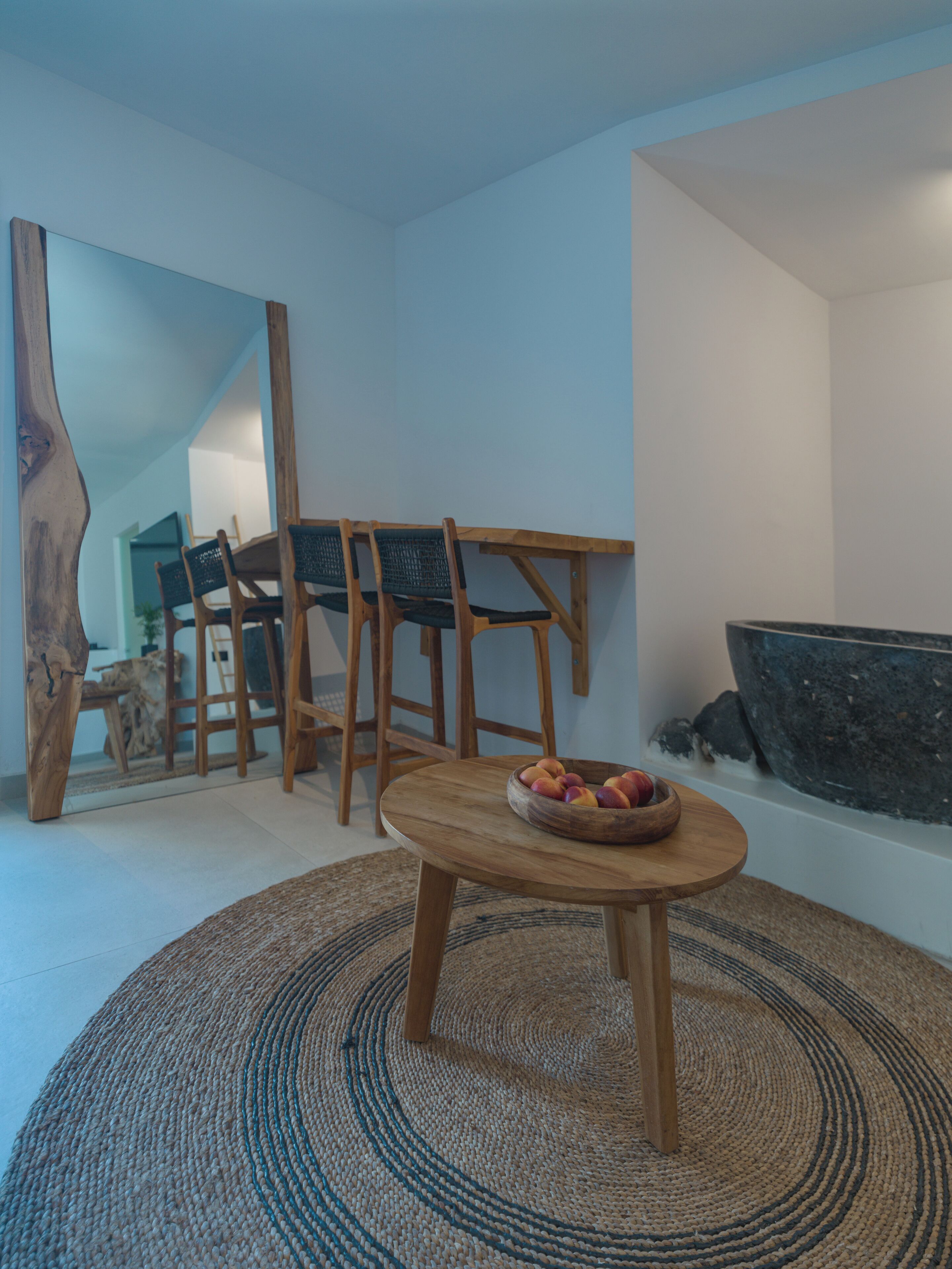 Uma sala de jantar moderna com uma mesa redonda de madeira, uma cesta de frutas e um espelho grande que reflete o ambiente.