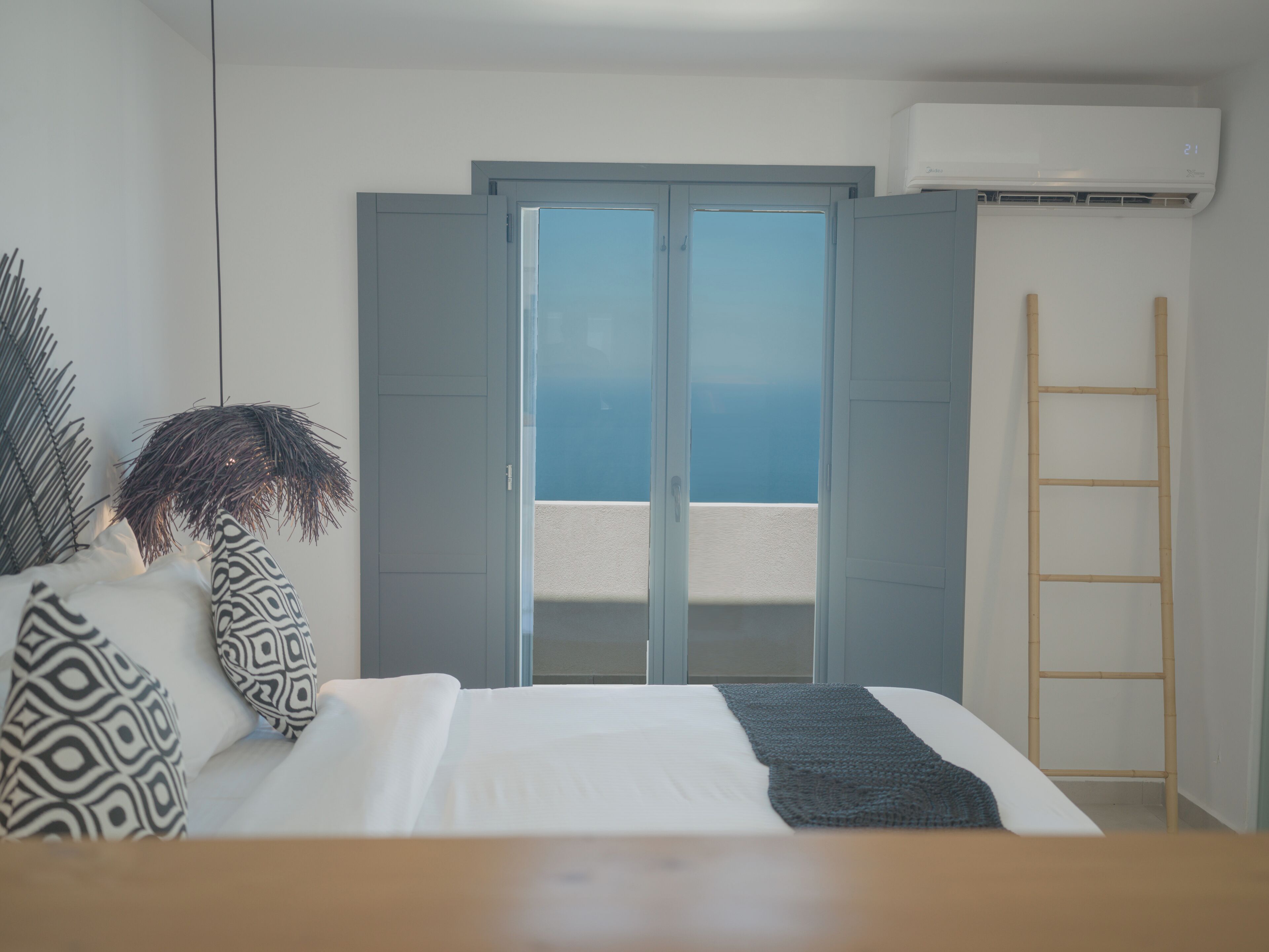 Quarto com cama, vista para o mar através de uma porta deslizante, escada e aparelho de ar condicionado.